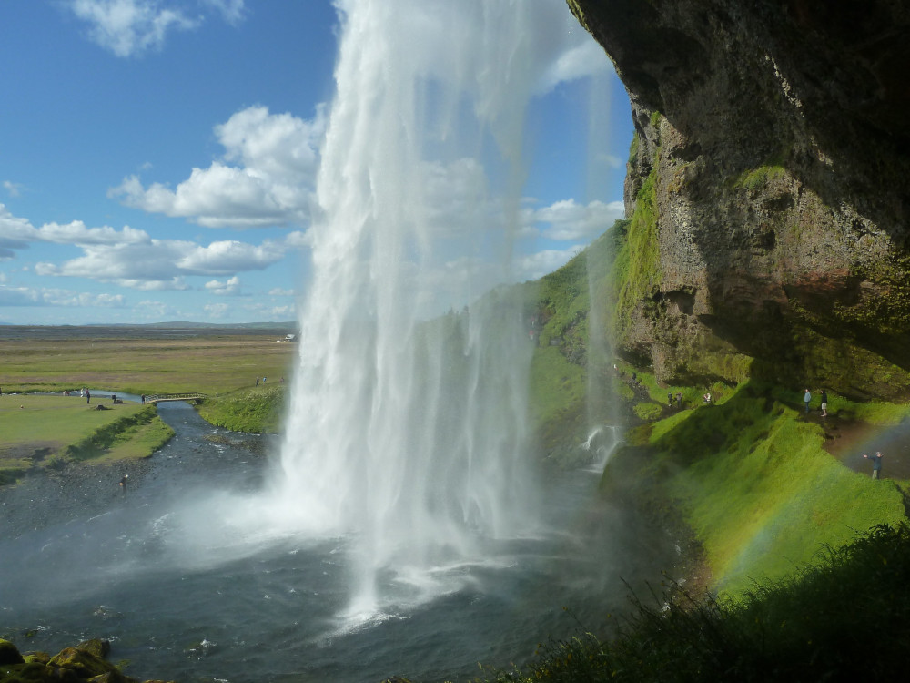 A tall slender lacy waterfall falls over the edge of a large cave into ...